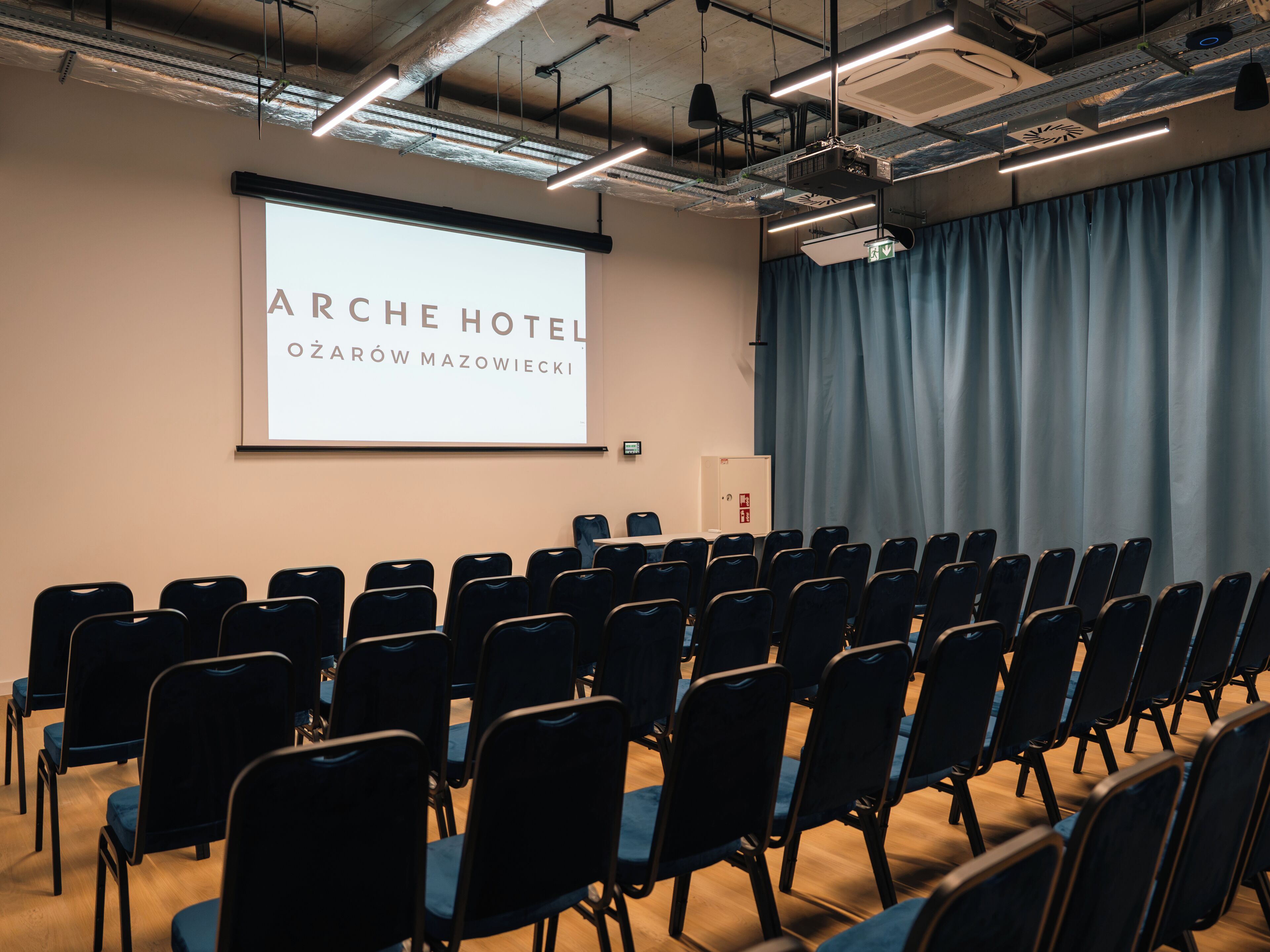 Uma sala de conferências com fileiras de cadeiras, uma tela de projeção exibindo 'ARCHE HOTEL Ozarów Mazowiecki' e uma cortina à direita.