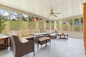 Um alpendre coberto com sofá, mesa de jantar e cadeiras, grandes janelas e um ventilador de teto.