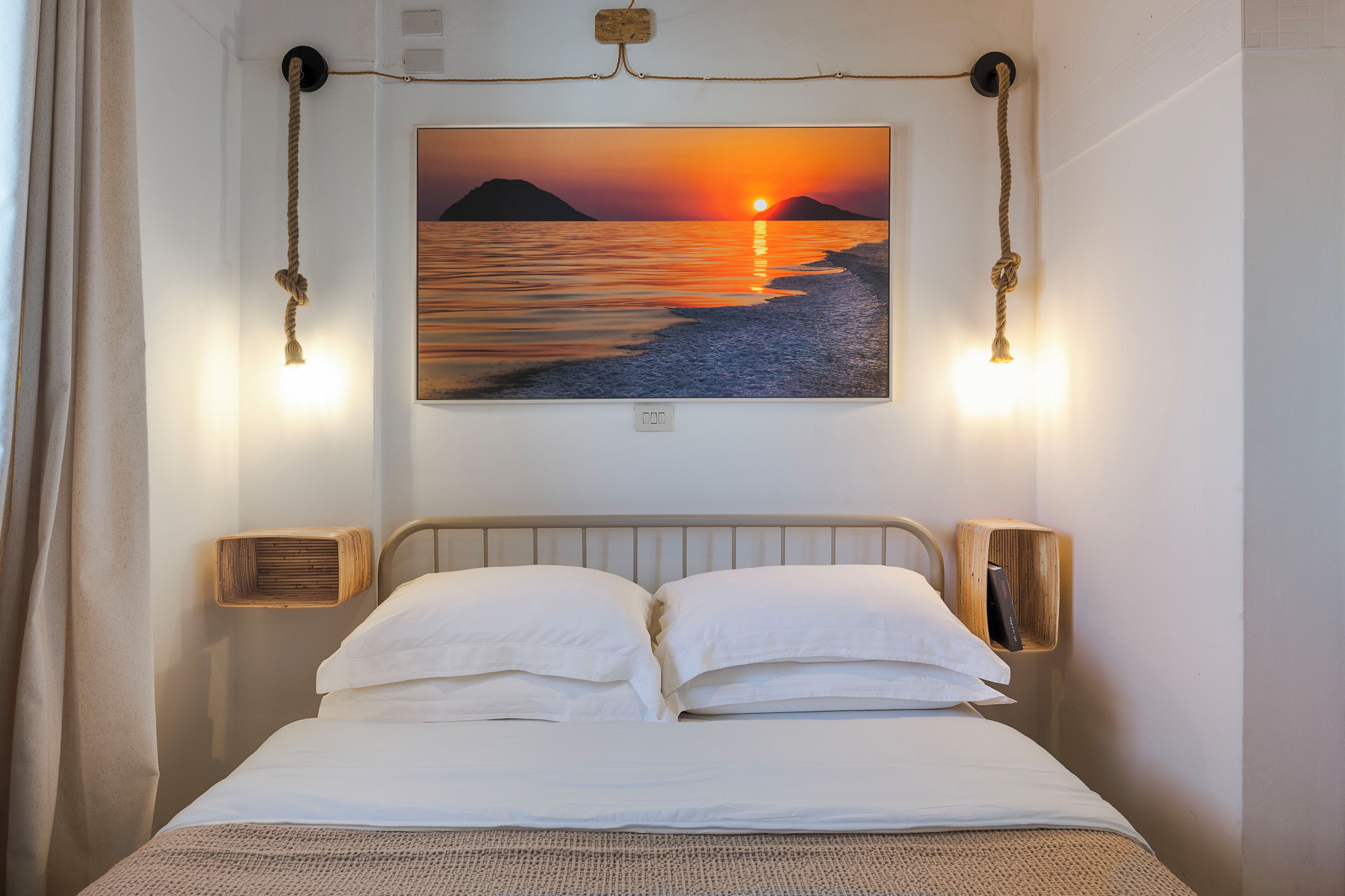 Uma cama com roupa de cama branca, um quadro com um pôr do sol sobre a água e duas colunas de som fixadas na parede.