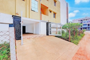 Um edifício residencial com vários andares, entrada com portão, área de estacionamento coberta e escadaria visível que dá acesso à entrada.