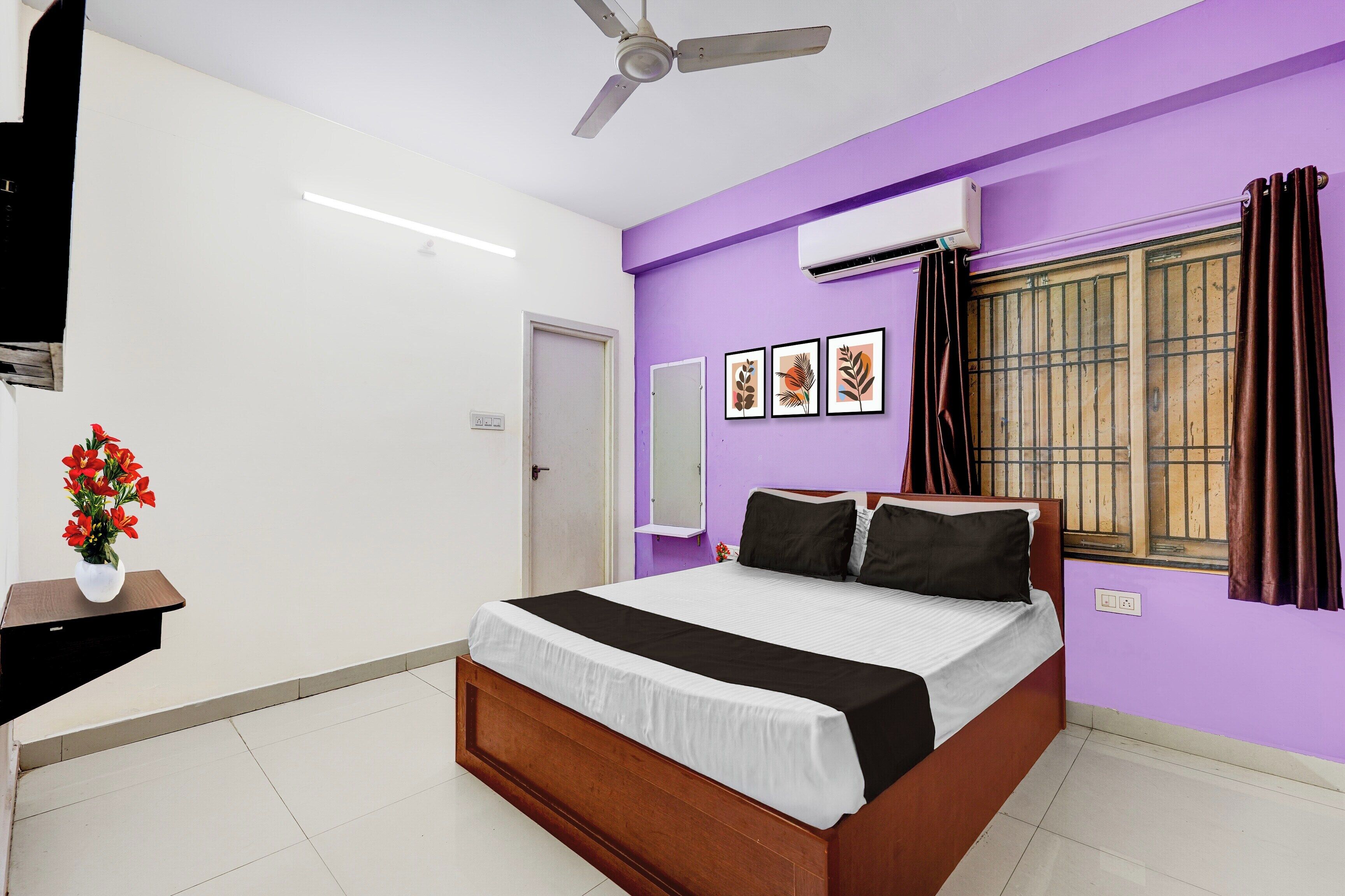 Quarto com cama, ventilador de teto, aparelho de ar condicionado, janela com cortinas e prateleira na parede com quadros.
