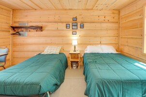 Quarto de cabana com duas camas, uma mesa pequena, uma cadeira e uma prateleira com fotografias.