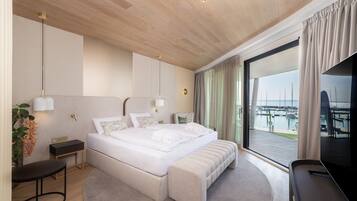 Quarto moderno com uma cama grande, mesas de cabeceira, televisão e vista para um porto de iates.