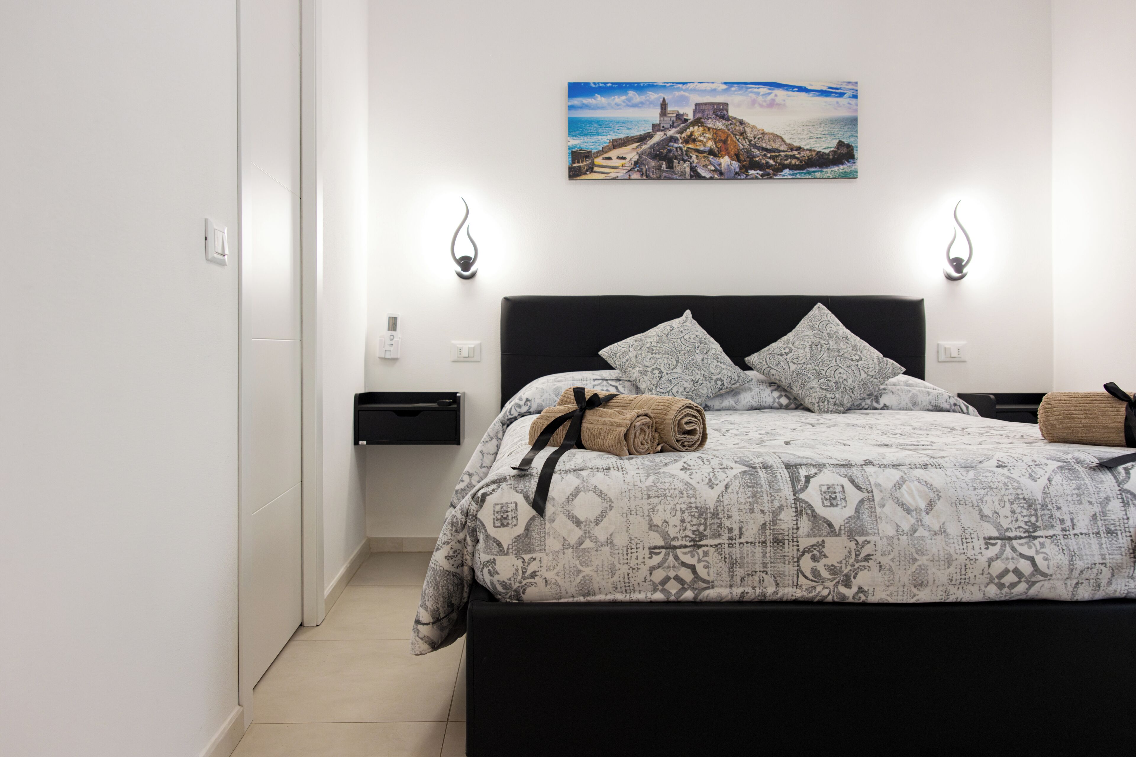 Quarto com cama, mesa de cabeceira, luminária na parede e uma pintura de uma paisagem costeira.