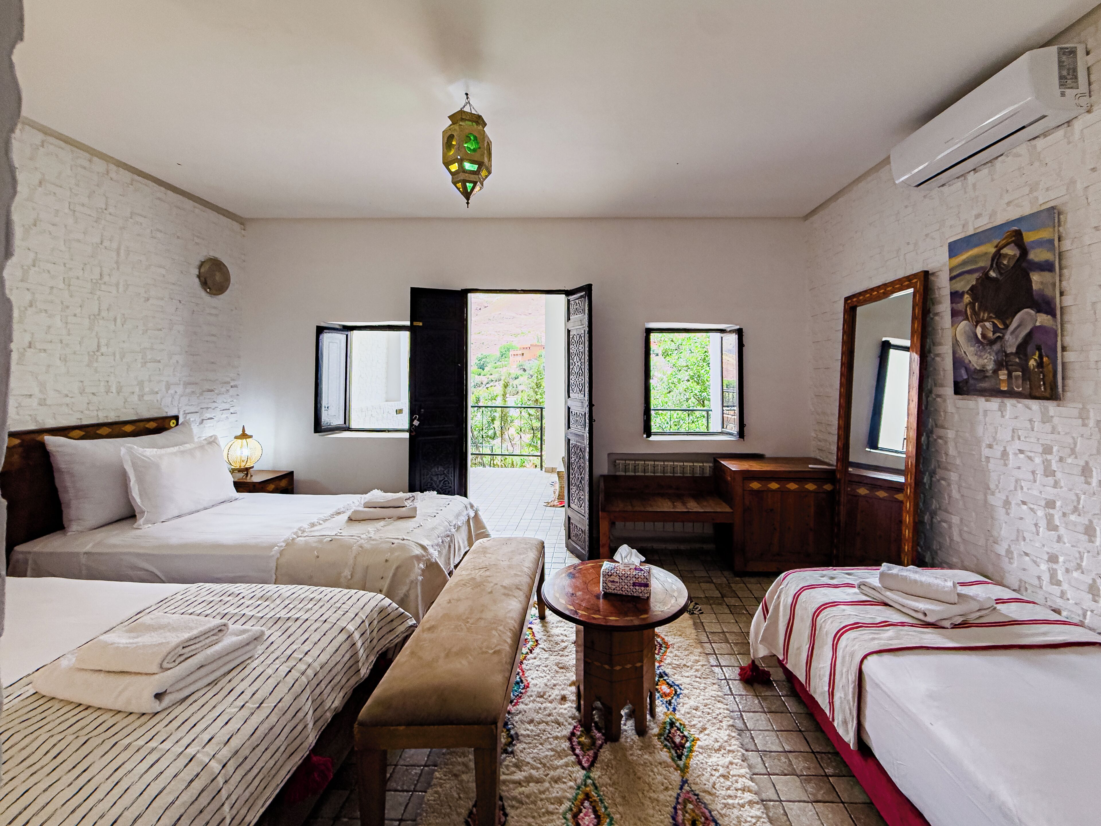 Quarto de hotel com duas camas, um banquinho, uma mesa pequena, um espelho e vista para o exterior.