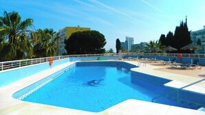 Piscina com deck azulejado, rodeada por palmeiras e edifícios.