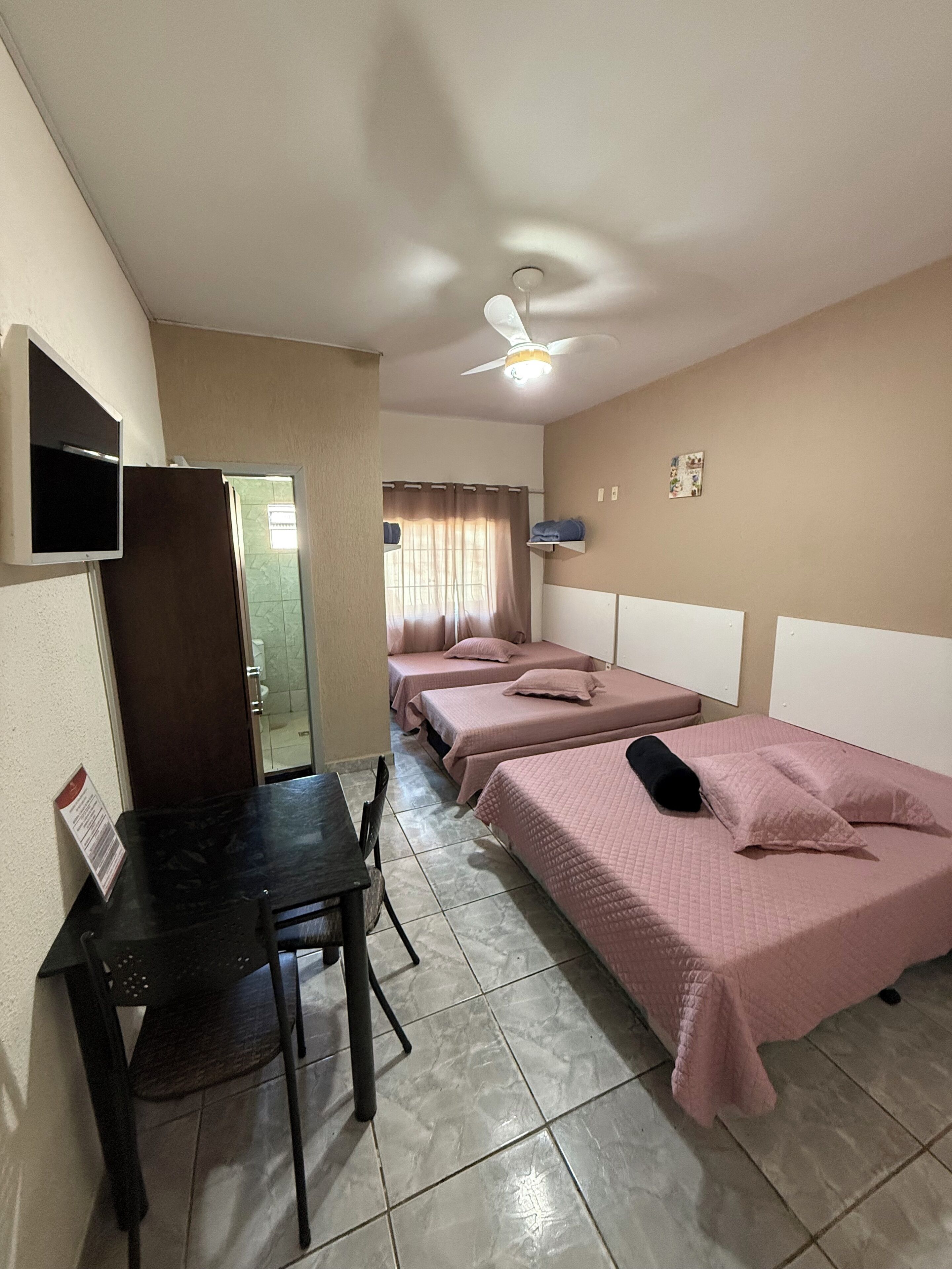 Quarto com duas camas, uma escrivaninha, uma cadeira, uma televisão e um ventilador de teto.