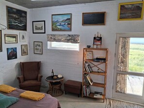 Um quarto com uma poltrona marrom, uma mesa redonda pequena com uma garrafa, uma estante e diversas obras de arte emolduradas nas paredes.