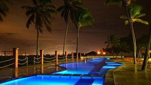 Piscina noturna com iluminação azul, cercada por palmeiras e uma grade.