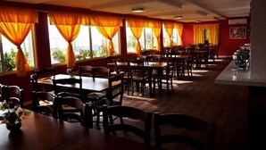 Restaurante com várias mesas e cadeiras, amplas janelas com cortinas laranja e vista para o exterior.