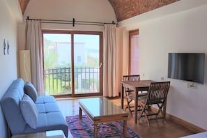 Uma sala de estar com sofá, mesa de centro de vidro, mesa de jantar com cadeiras, TV fixada na parede e porta de vidro deslizante que dá para uma área externa.