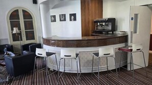 Um hall de hotel com um balcão de recepção curvo, banquetas de bar, uma máquina de café e um tapete estampado.