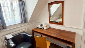 Quarto de hotel com escrivaninha, cadeira, radiador, telefone, espelho e janela com cortinas.