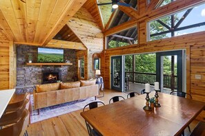 Interior acolhedor de uma cabana com lareira de pedra, um sofá, uma mesa de jantar e vista para o exterior.
