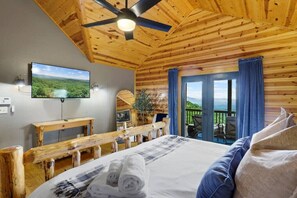 Quarto com cama, televisão, ventilador de teto, um banco e vista através de uma porta.