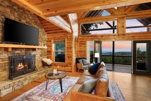 Interior acolhedor de uma cabana com lareira, televisão de tela plana, sofá e varanda com vista panorâmica.