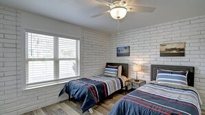 Quarto com duas camas, ventilador de teto, janela com persianas e um quadro na parede.
