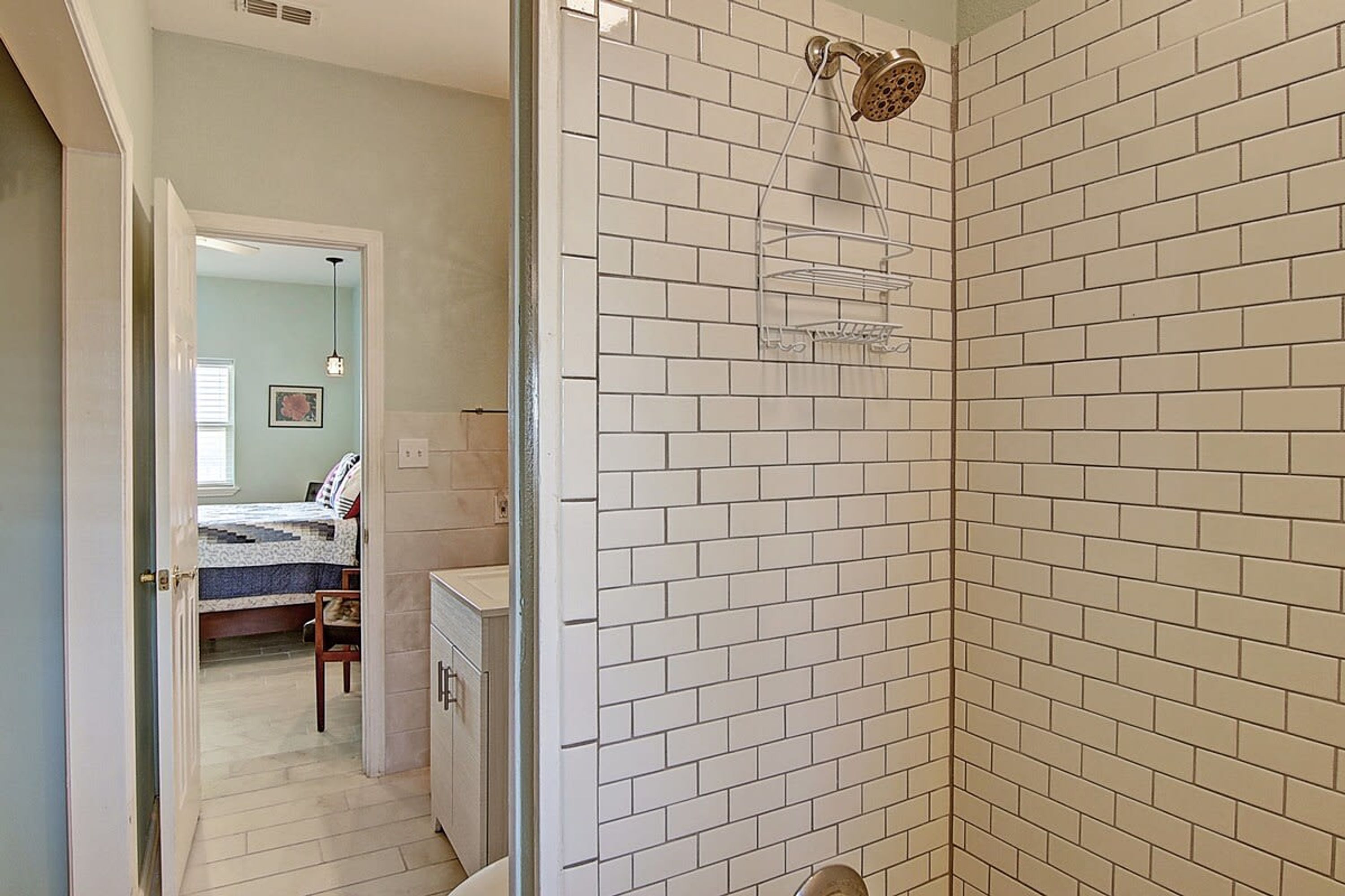 Um banheiro com azulejos brancos tipo metrô, chuveiro com barra e porta visível para um quarto com cama e poltrona.