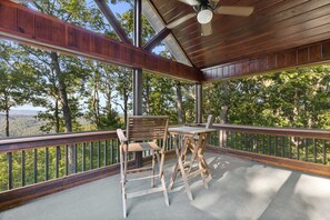 Uma área externa coberta com mesa e cadeiras, cercada por um corrimão de madeira, com vista para uma paisagem arborizada.