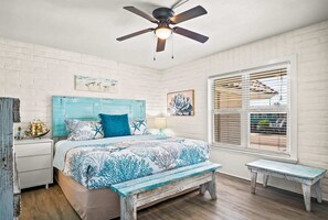 Quarto com cama, mesa de cabeceira, ventoinha no teto, janela com persianas e um banco.