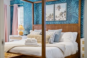 Uma cama de dossel com lençóis brancos, um abajur e quadros de parede com tema tropical.