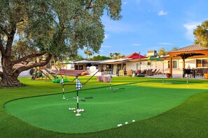 Um campo de golfe com putting green, uma rede para descanso e uma casa ao fundo.