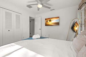 Um quarto com uma cama, um ventilador de teto, um quadro de um pôr do sol e uma porta.