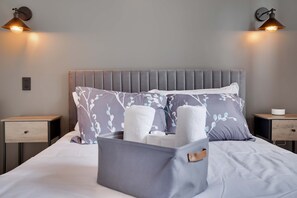 Cama bem arrumada com cabeceira cinza, duas mesas de cabeceira com luminárias e uma gaveta lateral.