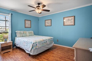 Um quarto com cama, mesa de cabeceira, ventilador de teto e quadros nas paredes.