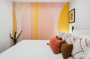 Quarto com cama, almofadas, uma planta de cacto e uma parede com arte abstrata.