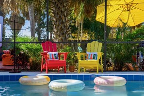 Uma área de piscina com duas cadeiras Adirondack, uma vermelha e outra amarela, ambas com almofadas 'RELAX'. Três boias infláveis estão na piscina.