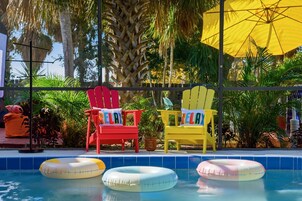 Uma área de piscina com duas cadeiras Adirondack, uma vermelha e outra amarela, ambas com almofadas 'RELAX'. Três boias infláveis estão na piscina.