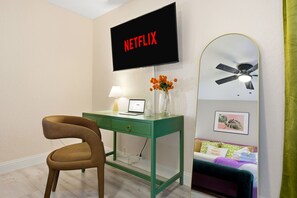 Um quarto com uma escrivaninha verde, uma cadeira marrom, uma TV fixada na parede, um espelho e um ventilador de teto.