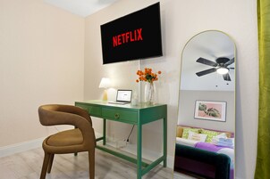Um quarto com uma escrivaninha verde, uma cadeira marrom, uma TV fixada na parede, um espelho e um ventilador de teto.