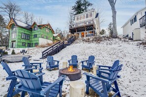 Uma área externa coberta de neve com cadeiras Adirondack ao redor de uma lareira, uma escadaria levando a um edifício e uma estufa em construção.