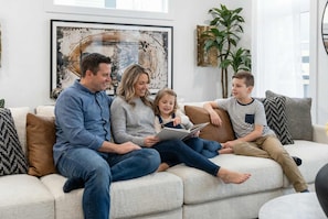 Uma família sentada em um sofá numa sala de estar com um quadro grande na parede e uma planta no canto.