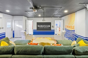 Uma sala de conferências com uma tela grande exibindo 'hometeam LUXURY RENTALS', um projetor e um sofá com almofadas.