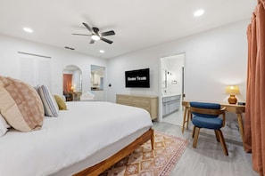 Quarto de hotel moderno com cama, escrivaninha, cadeira, televisão e ventoinha no teto.