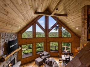 Uma sala de estar ampla com lareira de pedra, uma televisão e uma grande janela com vista para uma área arborizada.