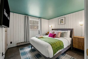 Um quarto com cama, mesa de cabeceira, televisão na parede, janela com cortinas e luminária de parede.