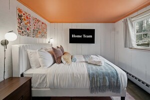 Quarto com cama, almofadas, mesa de cabeceira, televisão fixada na parede e janela com cortinas.
