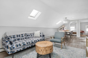 Um loft moderno com pé direito alto, uma mesa de jantar de madeira, um sofá azul com almofadas estampadas e um banquinho de madeira redondo.