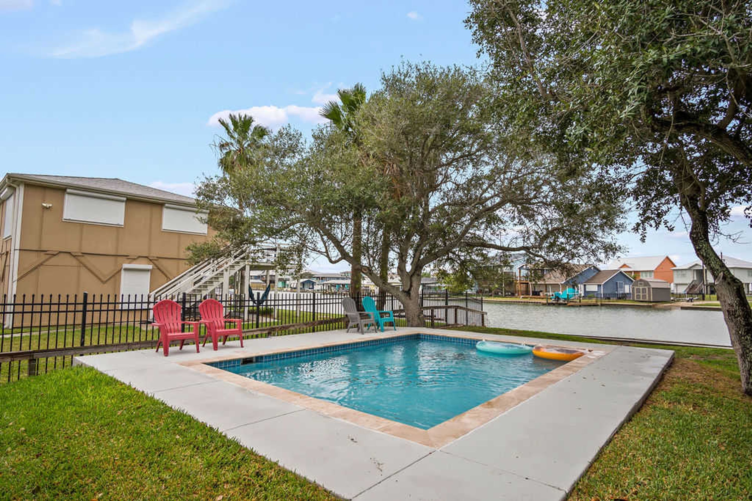 Uma piscina retangular com borda azulejada, rodeada por um deck de concreto. Há duas cadeiras Adirondack vermelhas e um bote inflável azul.