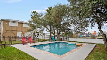 Uma piscina retangular com borda azulejada, rodeada por um deck de concreto. Há duas cadeiras Adirondack vermelhas e um bote inflável azul.