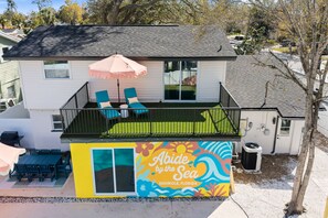 Uma casa de dois andares com uma varanda, um pátio com cadeiras e um guarda-sol, e um mural colorido na lateral.