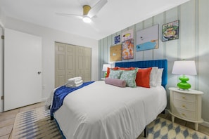 Quarto de hotel com cama, mesa de cabeceira, ventilador de teto e obras de arte na parede.
