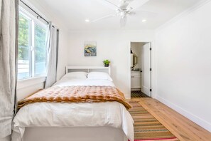 Quarto com cama, ventilador de teto, janela com cortinas, porta para um banheiro e uma pequena planta na mesa de cabeceira.