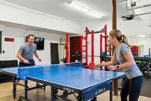 Mesa de pingue-pongue com duas pessoas jogando, cercada por equipamentos de ginástica e um relógio digital.
