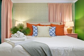 Quarto de hotel com cama, almofadas, mesa de cabeceira, abajur, planta e uma pilha de toalhas.
