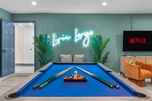 Uma mesa de bilhar com bolas e tacos, um letreiro de neon que diz 'Lurin Large', uma televisão exibindo 'NETFLIX' e uma porta que dá para outro cômodo.